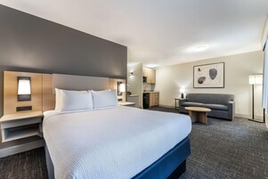 Quarto de hotel com uma cama grande, um sofá, uma mesinha e uma cozinha compacta.