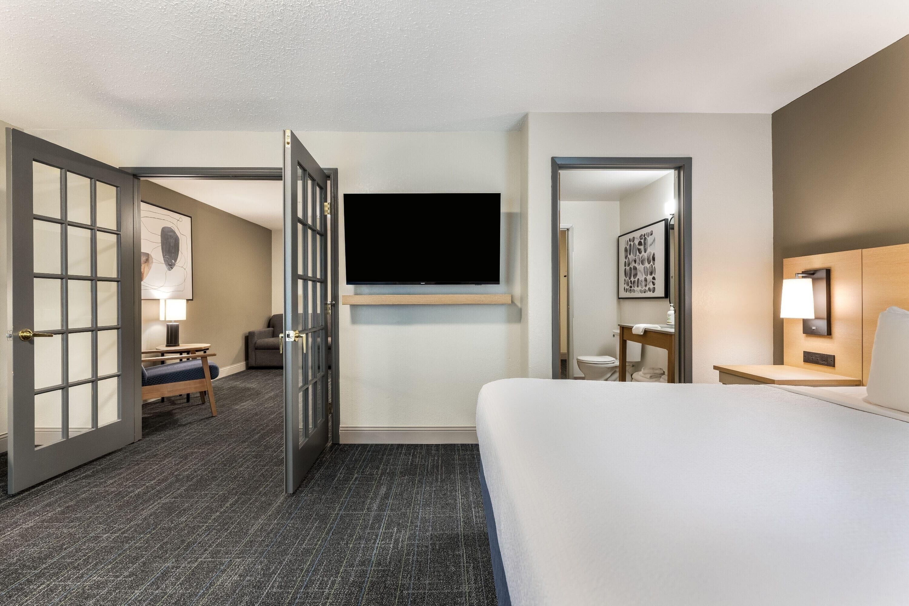Quarto de hotel com cama, escrivaninha, cadeira, televisão e banheiro.