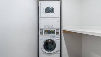Uma máquina de lavar e secar empilhadas em uma lavanderia, com uma parede branca e uma prateleira de madeira acima.