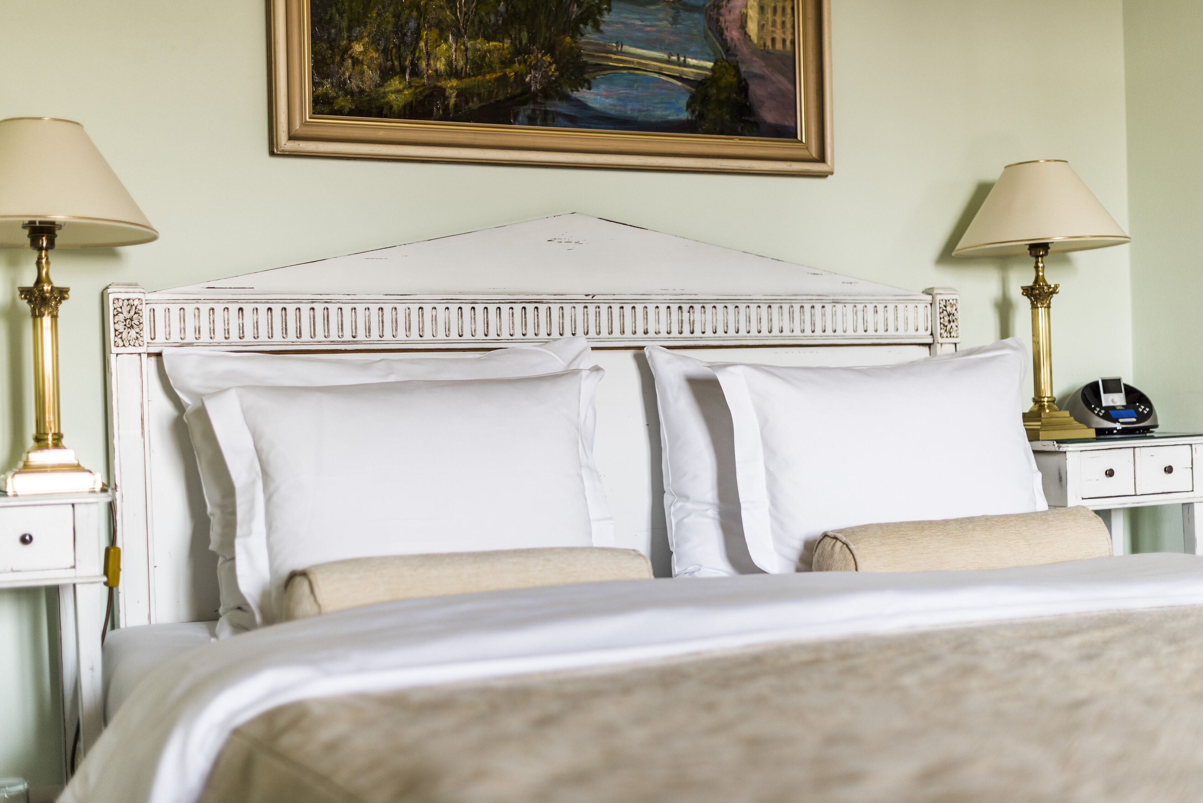Cama bem arrumada com roupa de cama branca e cabeceira com molduras decorativas. Duas mesas de cabeceira, uma delas com luminária e um pequeno relógio. Um quadro emoldurado está pendurado acima da cama.