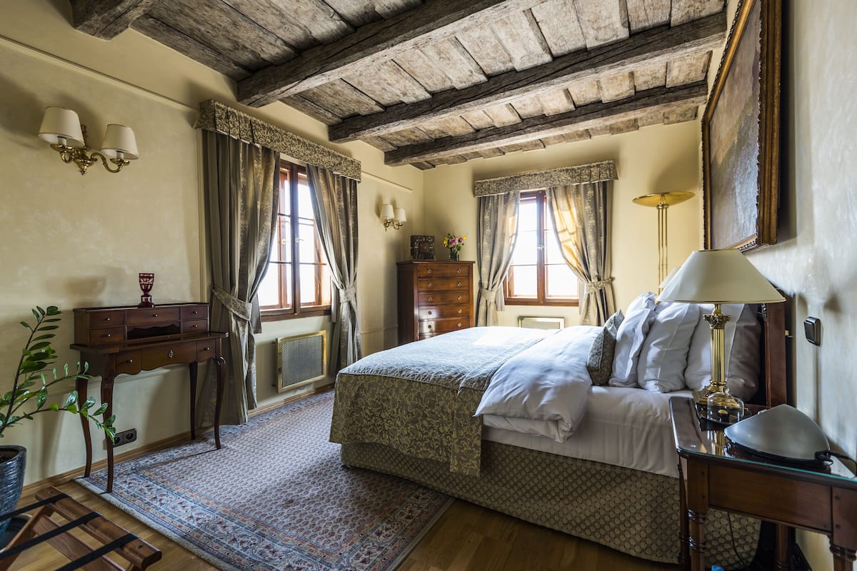 Quarto com teto de madeira, cama com lençóis brancos, mesa de cabeceira com abajur, cômoda e janela com cortinas.