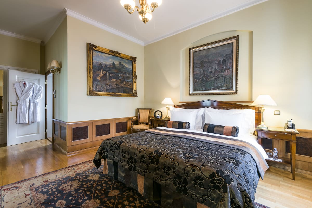Quarto de hotel com uma cama grande, duas pinturas nas paredes, uma mesinha de cabeceira de madeira, uma escrivaninha e um porta-robes.