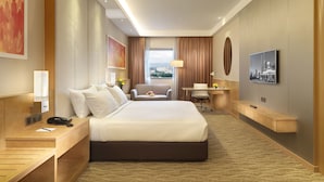 Quarto de hotel com uma cama grande, uma escrivaninha com cadeira, uma mesinha de apoio com abajur e uma televisão na parede.