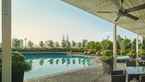 Área da piscina com espreguiçadeiras e mesas, vista para o horizonte da cidade e um terraço coberto com ventiladores de teto.