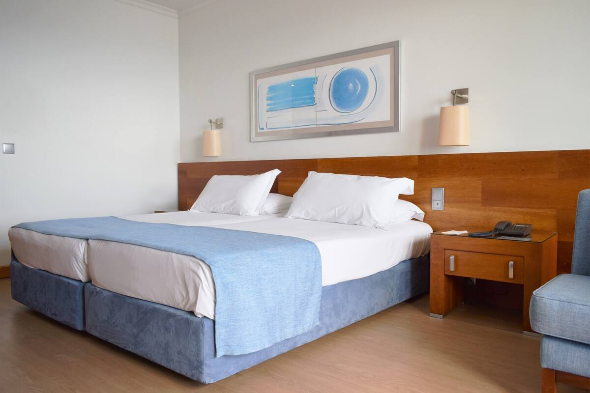 Quarto de hotel com uma cama grande, cabeceira de madeira, mesa de cabeceira com abajur e um quadro na parede.