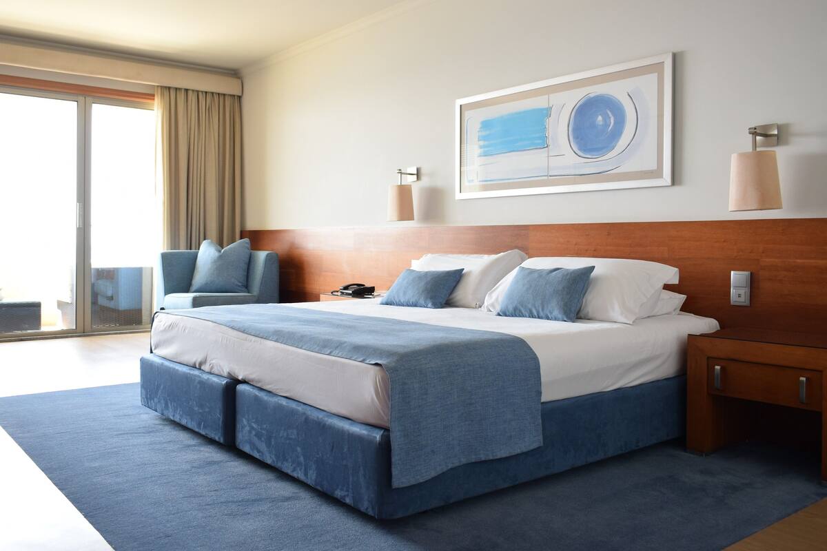 Quarto de hotel moderno com uma cama grande, cabeceira de madeira, uma janela ampla com cortinas, um quadro na parede e uma mesinha de cabeceira com abajur.