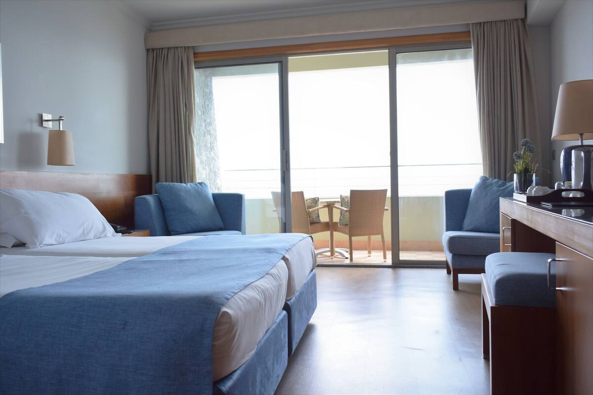 Quarto de hotel com uma cama grande, uma escrivaninha, uma cadeira e varanda com mesa e cadeiras.