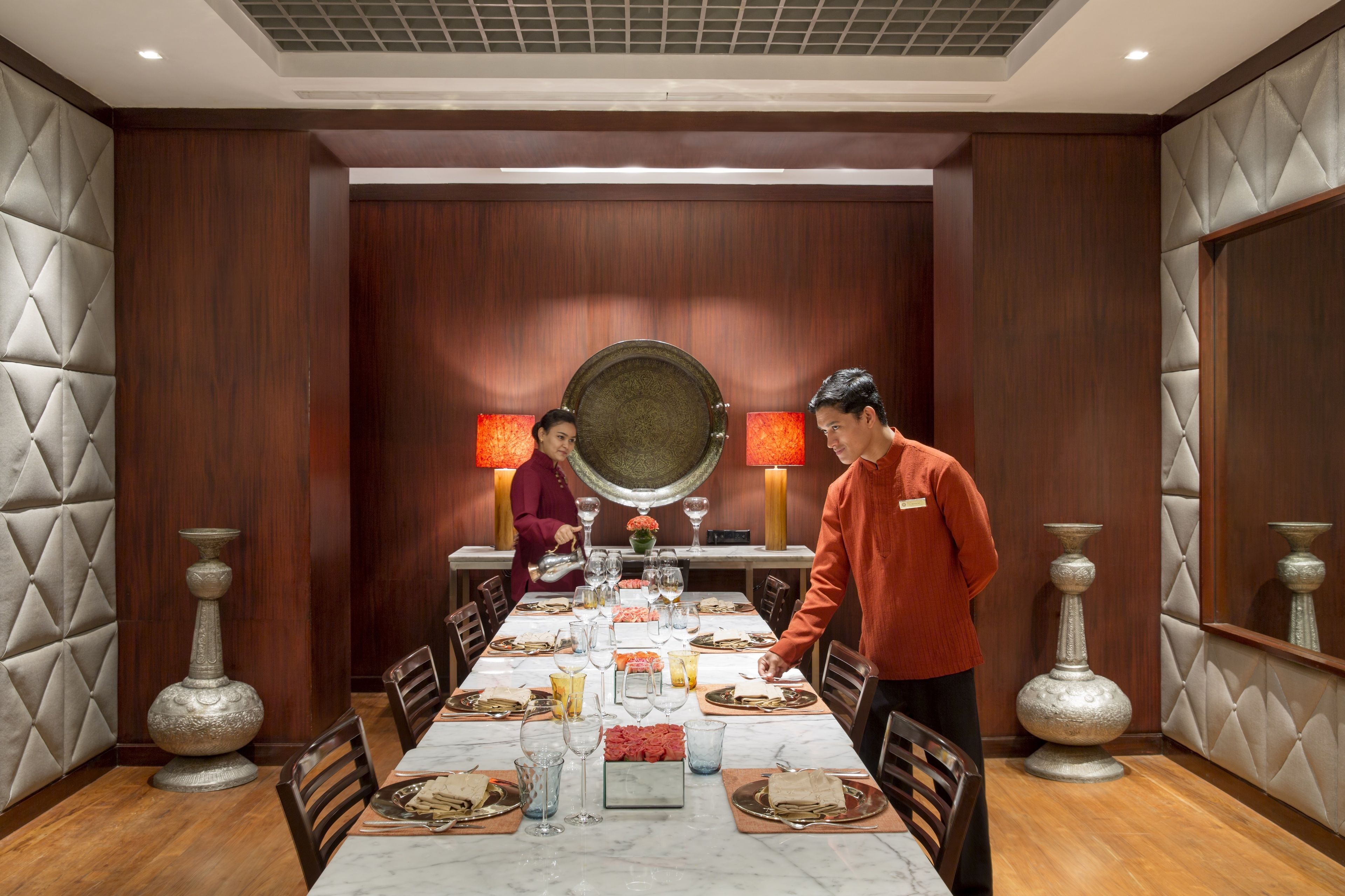 Uma sala de jantar formal com uma mesa longa posta para refeição, dois funcionários e painéis decorativos nas paredes.