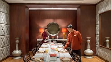 Uma sala de jantar formal com uma mesa longa posta para refeição, dois funcionários e painéis decorativos nas paredes.