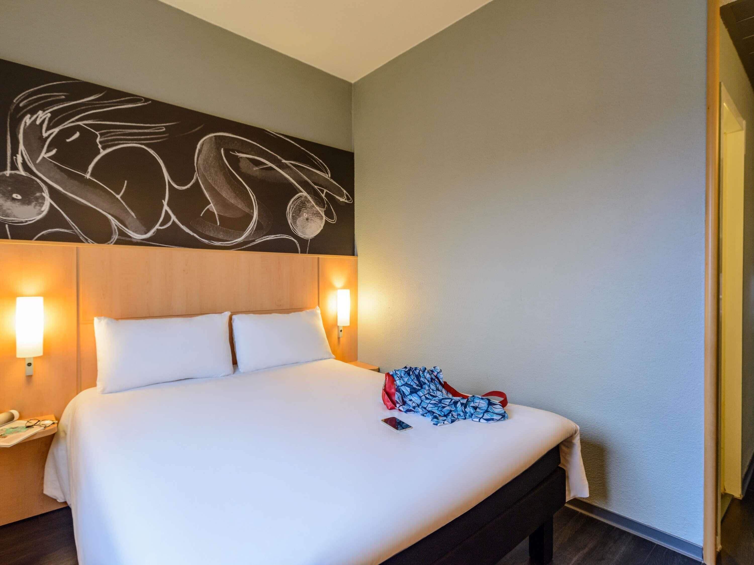 Quarto de hotel com cama grande, duas luminárias de cabeceira, uma obra de arte na parede e uma mala sobre a cama.