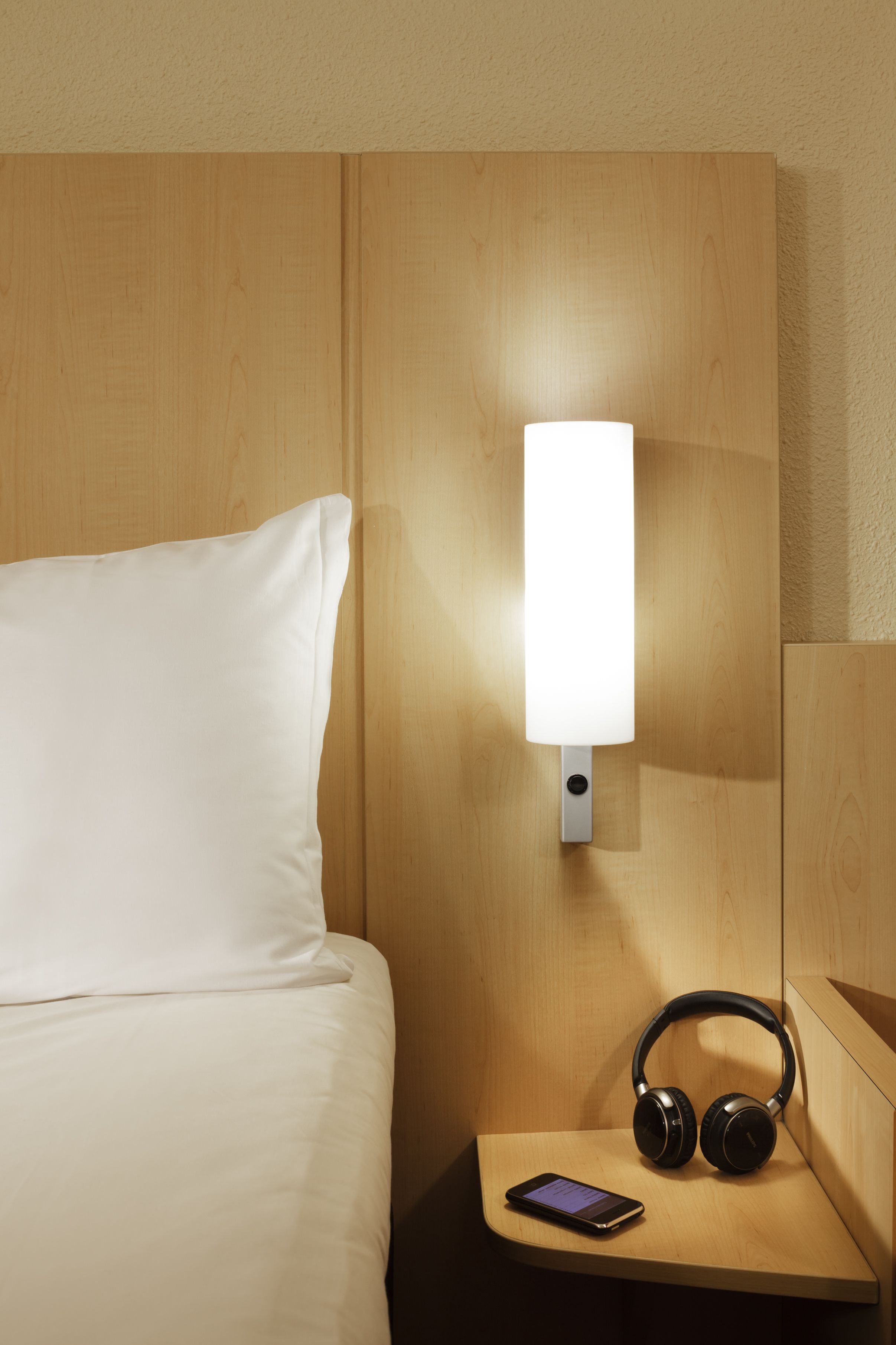 Uma cama bem arrumada com lençóis brancos, uma mesinha de cabeceira com um smartphone e fones de ouvido, e um ponto de luz fixado na parede.