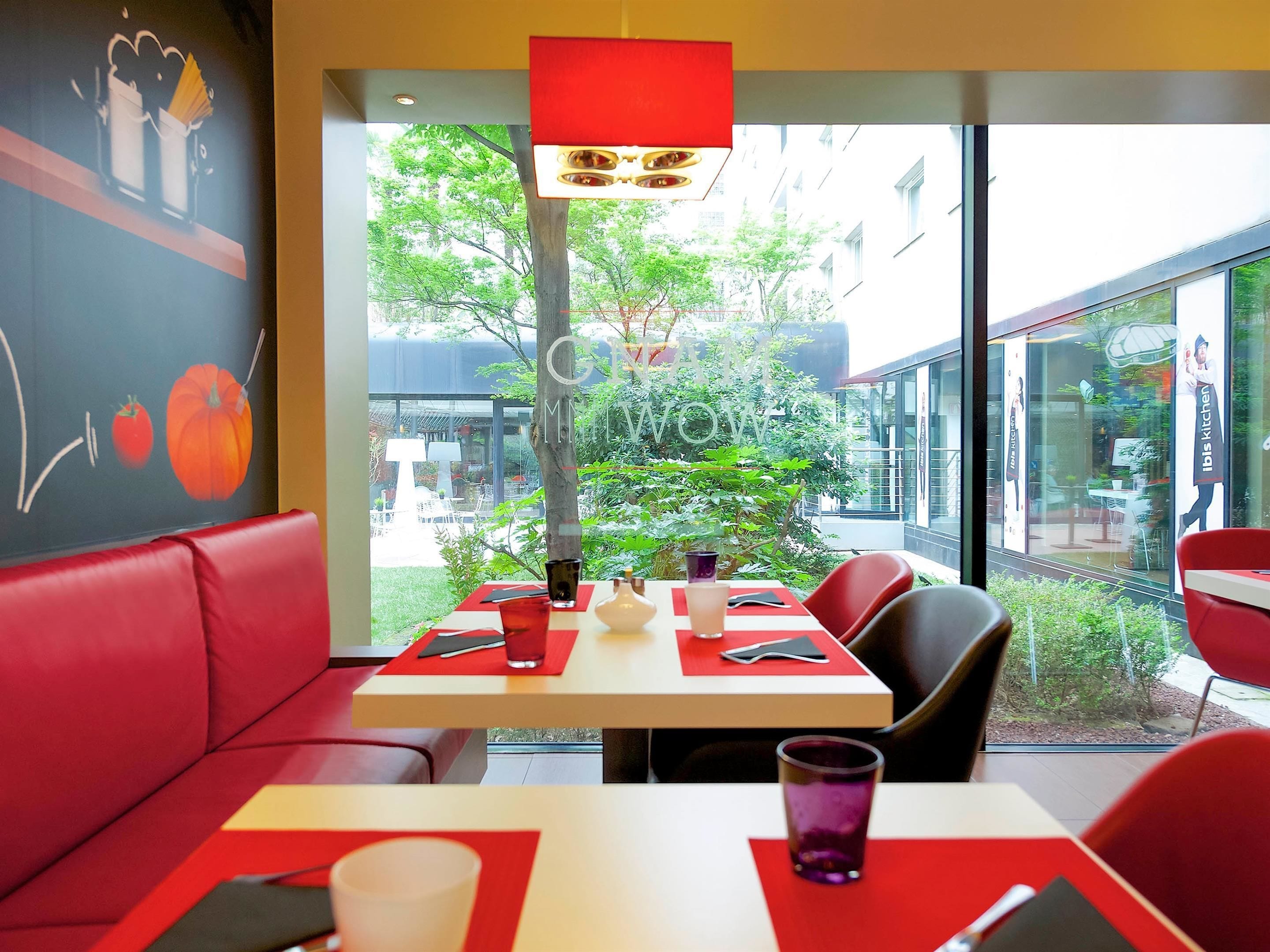 Restaurante com assentos vermelhos, mesa com jogo americano vermelho e vista para um jardim e edifícios lá fora.