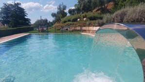 Uma piscina com vista clara da água, uma estrutura metálica à direita e uma área ajardinada com cadeiras e plantas ao fundo.