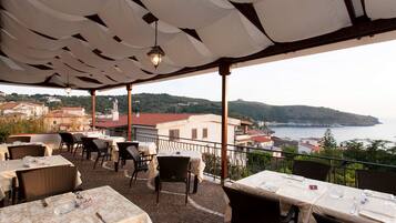 Terraço ao ar livre com mesas e cadeiras, com vista para uma cidade costeira e o mar.