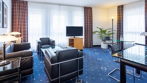 Quarto de hotel com sofá, poltrona, mesa de vidro, televisão e escrivaninha com luminária.