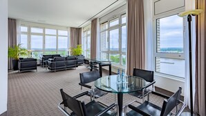 Quarto de hotel moderno com uma mesa de vidro grande, assentos confortáveis e vista da cidade por grandes janelas.