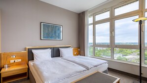 Quarto de hotel com uma cama grande, uma mesa de cabeceira, um abajur, um quadro na parede e vista para a cidade através de amplas janelas.