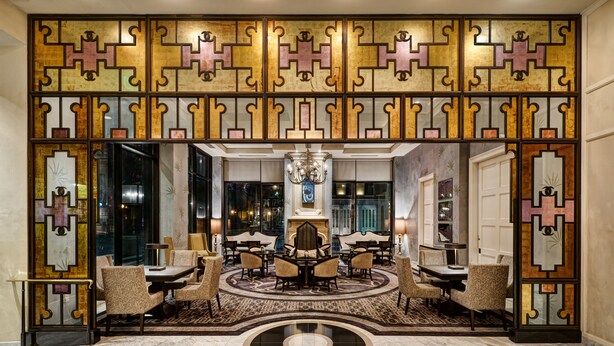 Um hall com área de estar central, um candelabro e uma parede decorativa com padrões geométricos.
