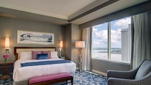 Quarto de hotel com uma cama grande, um banco, uma cadeira, vista da cidade e um quadro na parede.