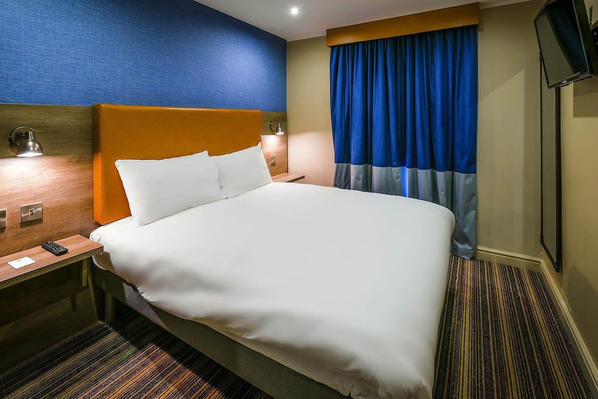 Quarto de hotel com uma cama grande, cabeceira de madeira, mesa de cabeceira com luminária, televisão fixada na parede e janela com cortinas azuis.
