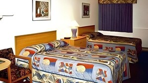 Quarto de hotel com duas camas, cabeceira de madeira, mesa de cabeceira com abajur e uma cadeira.