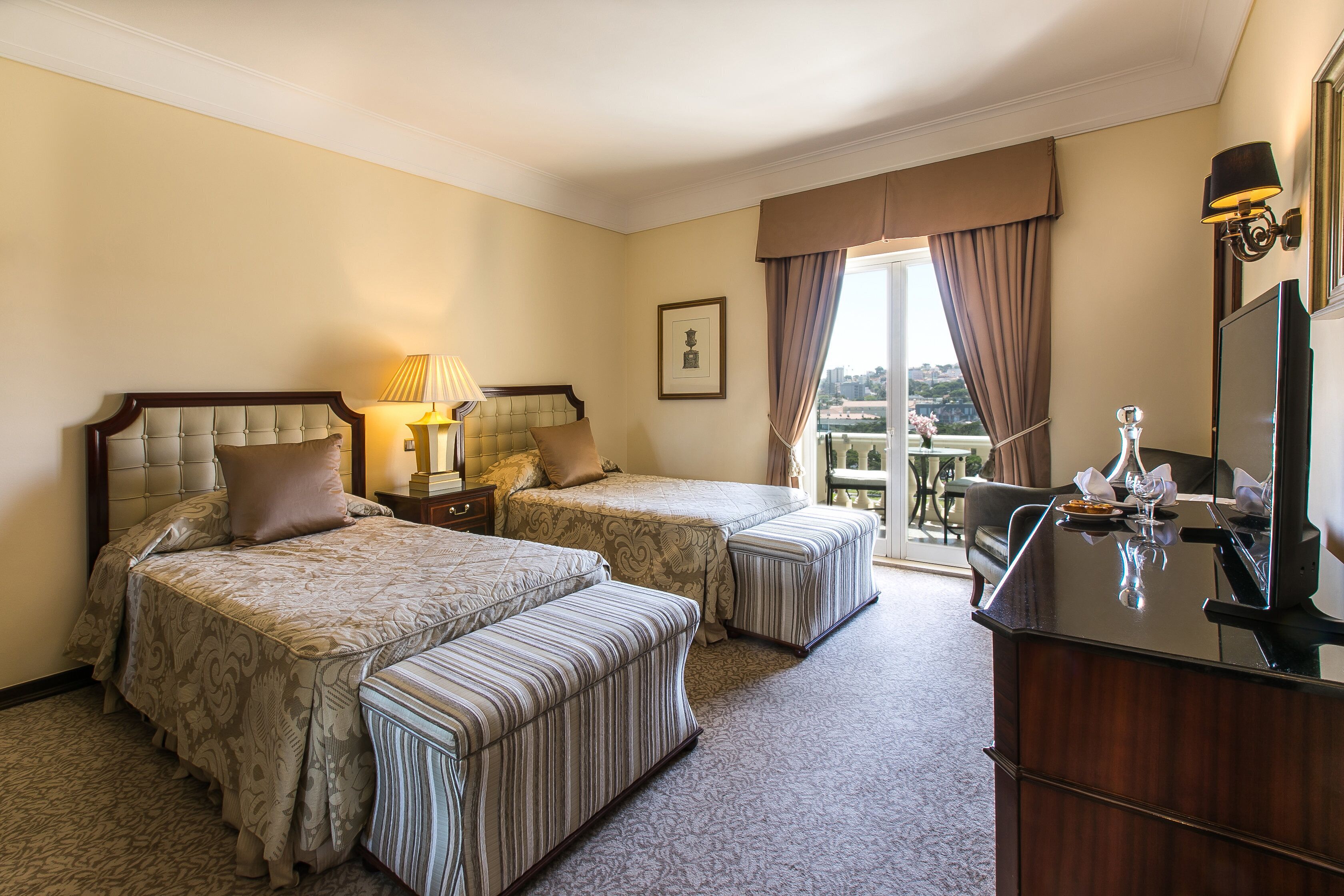 Quarto de hotel com duas camas, uma escrivaninha com espelho, cómoda e varanda com vista.