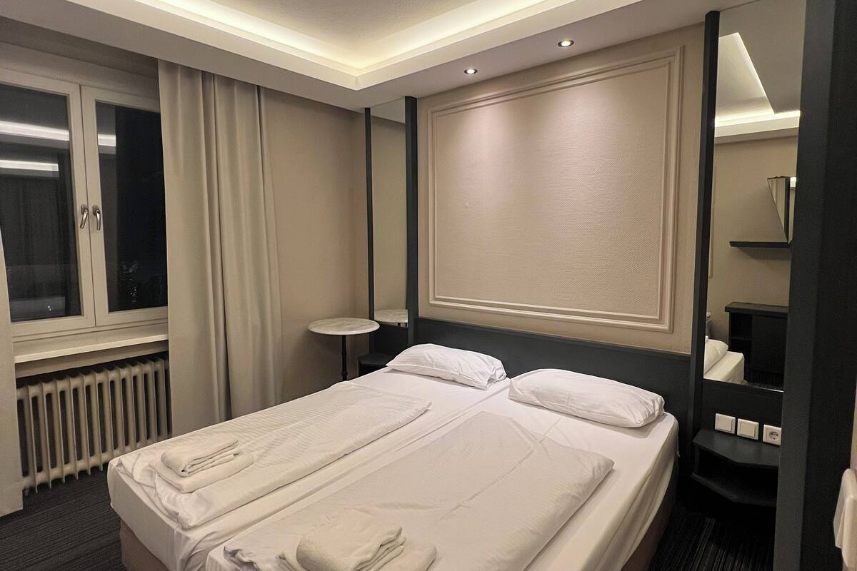Quarto de hotel com uma cama grande, uma mesa de cabeceira, um espelho e uma janela com cortinas.