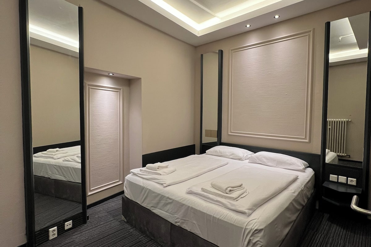 Quarto de hotel com uma cama grande, uma mesa de cabeceira, um espelho e um lustre no teto.