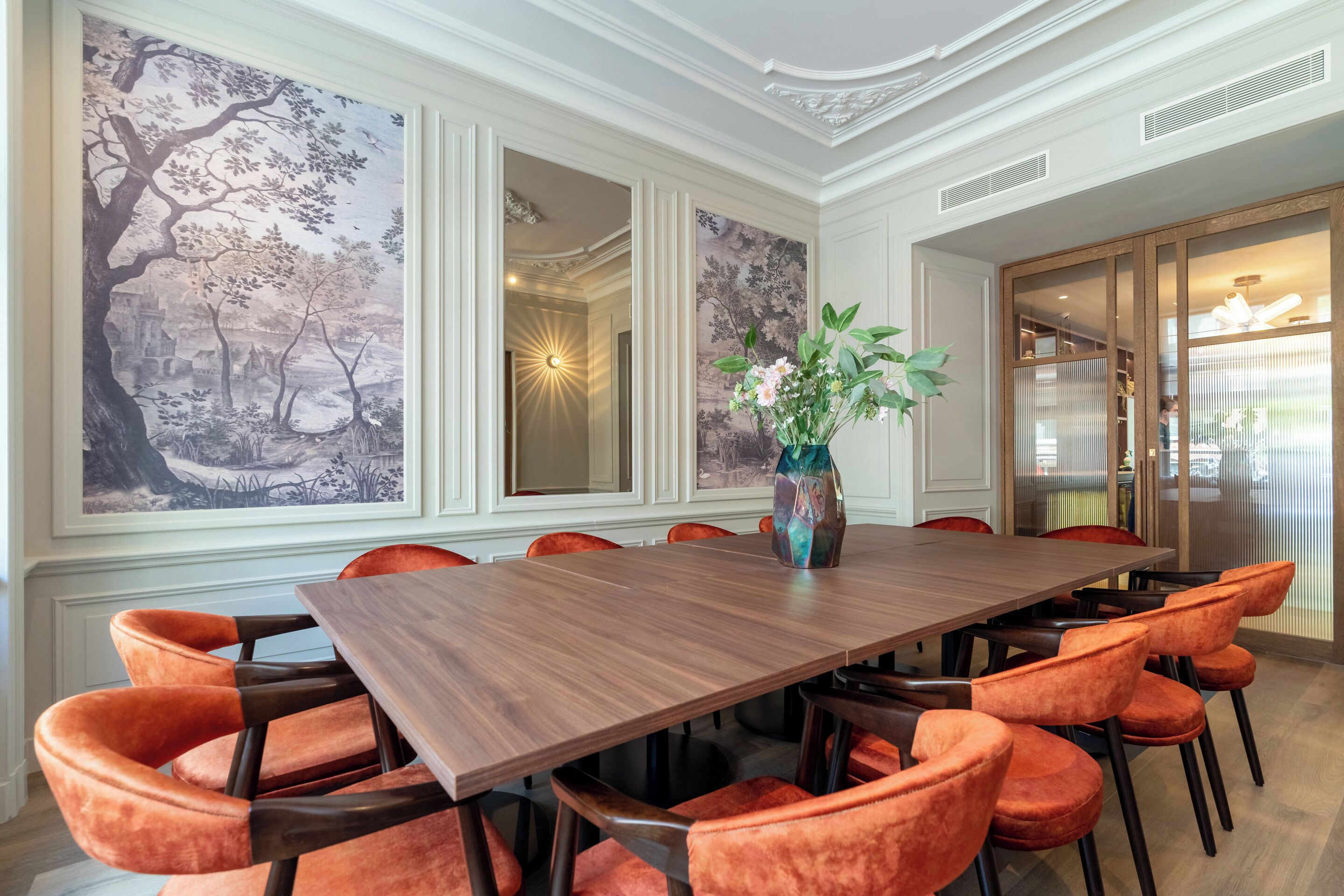 Uma sala de jantar com uma mesa de madeira comprida, cadeiras estofadas em laranja e grandes pinturas penduradas na parede.