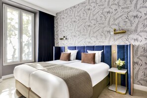 Uma cama de casal com colcha bege, cabeceira azul e uma pequena mesa de cabeceira com um vaso de flores.