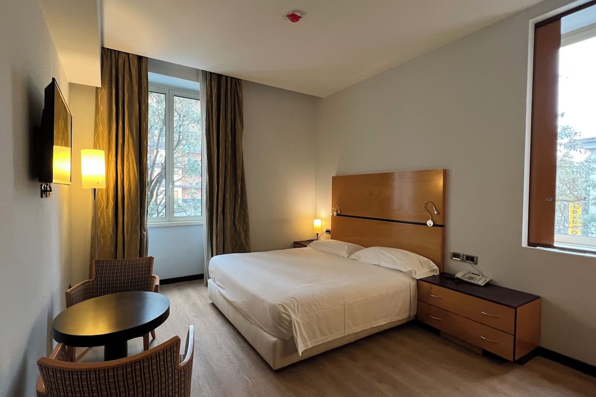 Quarto de hotel com cama, mesa de cabeceira, luminária, cadeira e janela com cortinas.