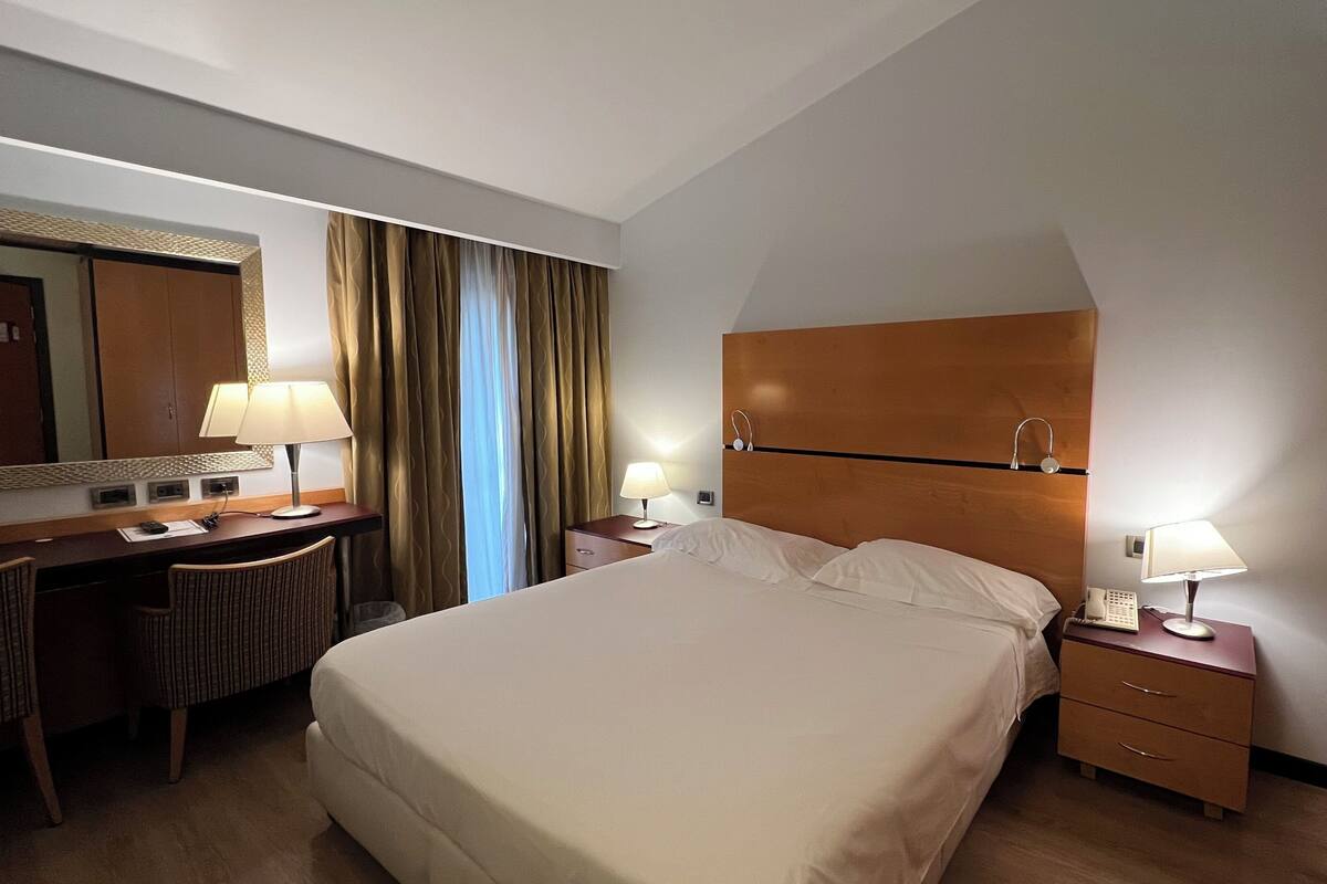 Quarto de hotel com cama, mesas de cabeceira, escrivaninha, cadeira, espelho e cortinas.