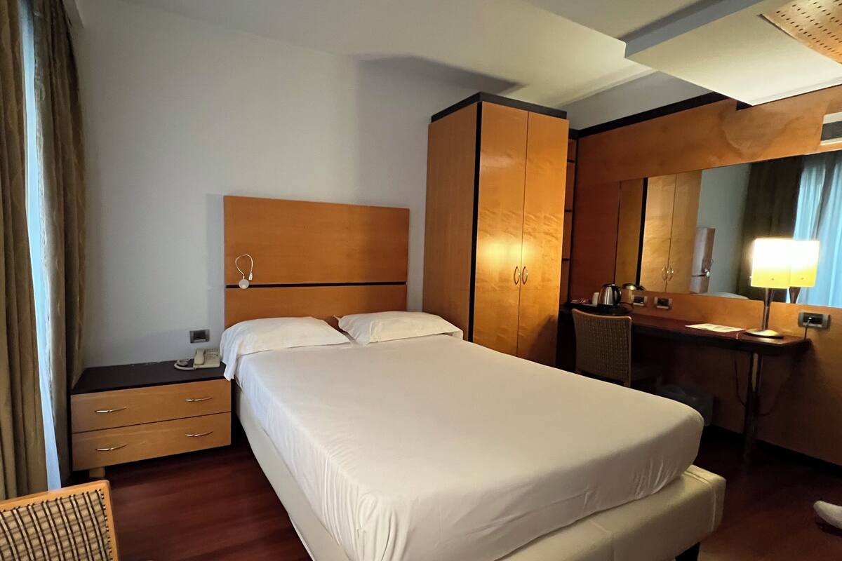 Quarto de hotel com cama, escrivaninha, cadeira, guarda-roupa e espelho.