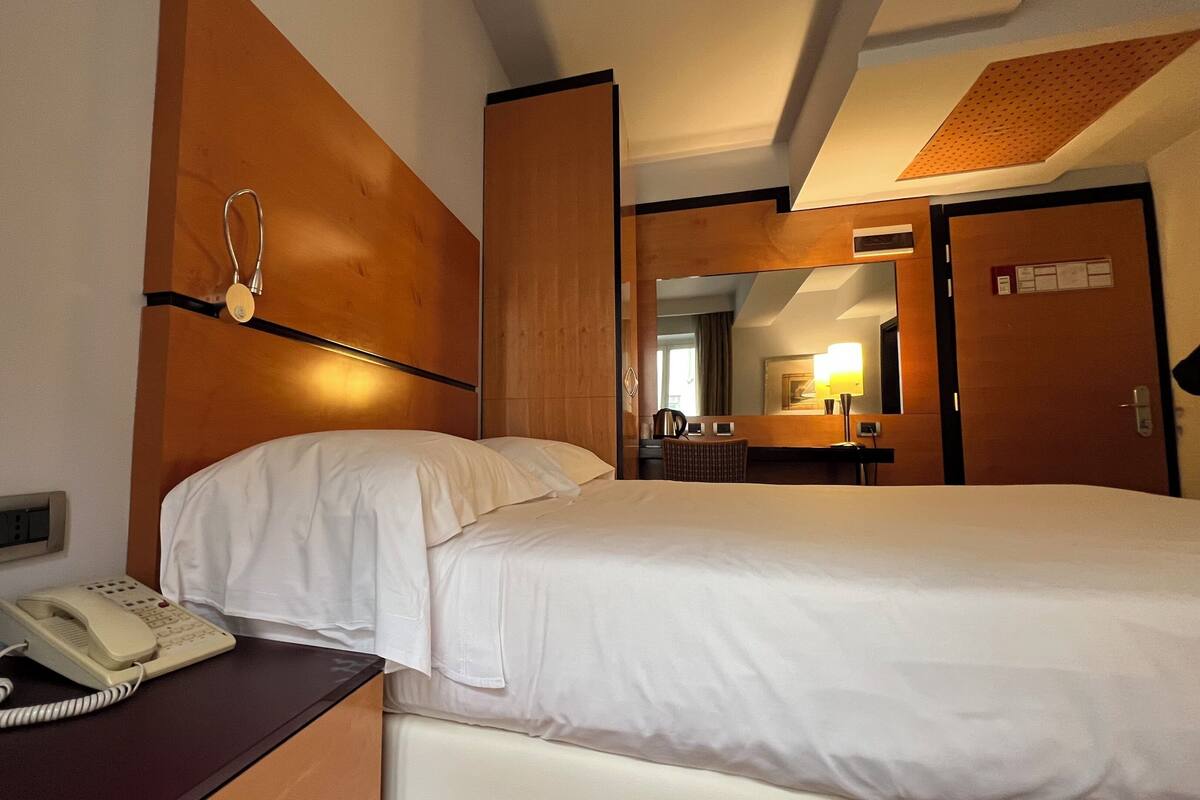 Quarto de hotel com uma cama, mesa de cabeceira com telefone, espelho e cabeceira de madeira.