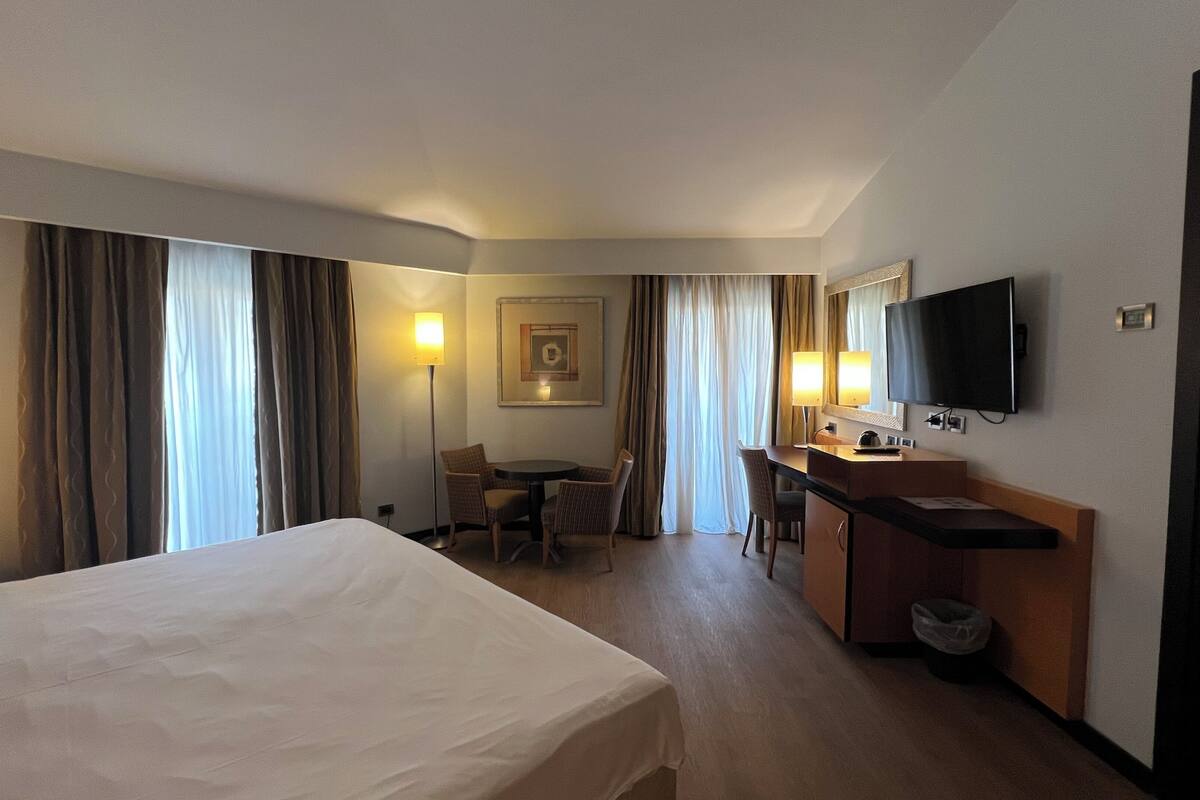 Quarto de hotel com cama, escrivaninha com cadeira, televisão e um quadro na parede.