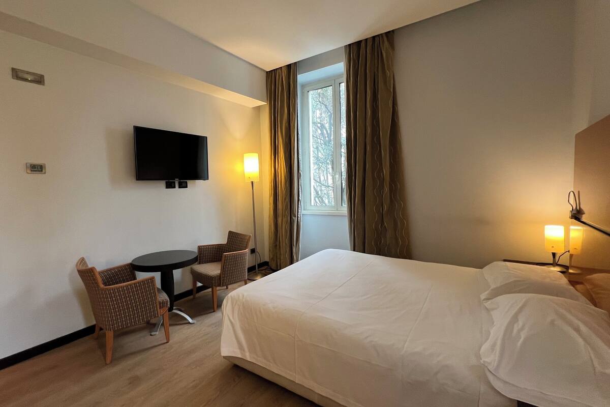 Quarto de hotel com cama, televisão na parede, escrivaninha com duas cadeiras e janela com cortinas.