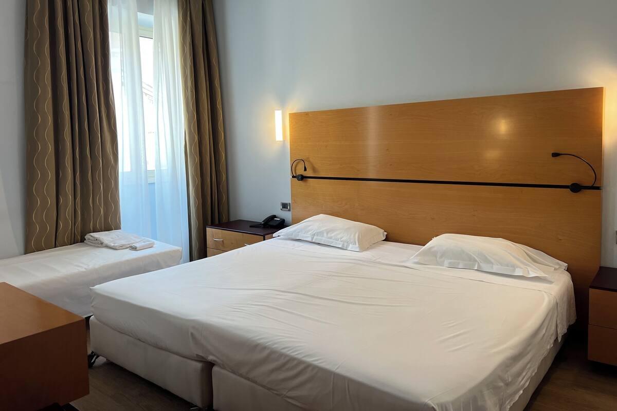 Quarto de hotel com uma cama grande, mesa de cabeceira e janela com cortinas.
