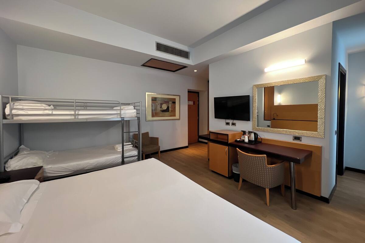 Quarto de hotel com beliches, escrivaninha, cadeira, televisão, espelho e um quadro na parede.