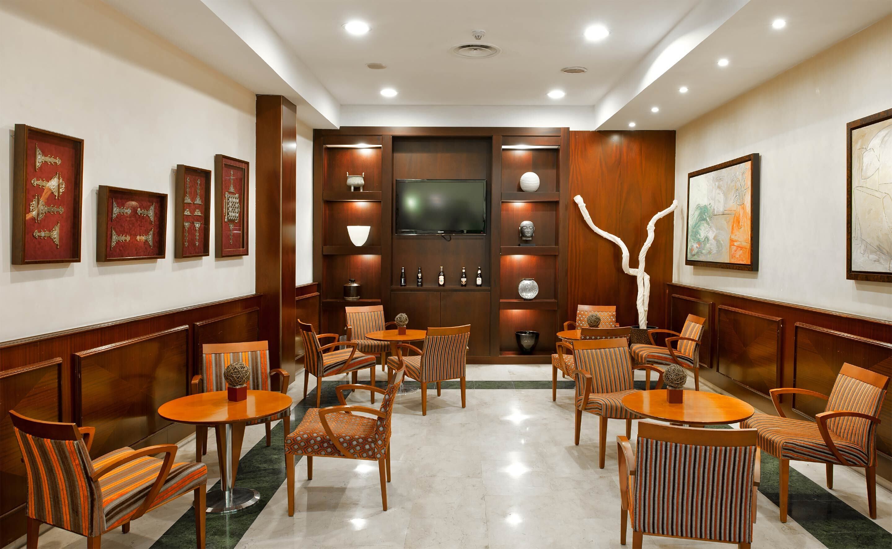 Um saguão de hotel com painéis de madeira, uma área de estar central com mesas redondas e cadeiras listradas, e uma televisão montada na parede.