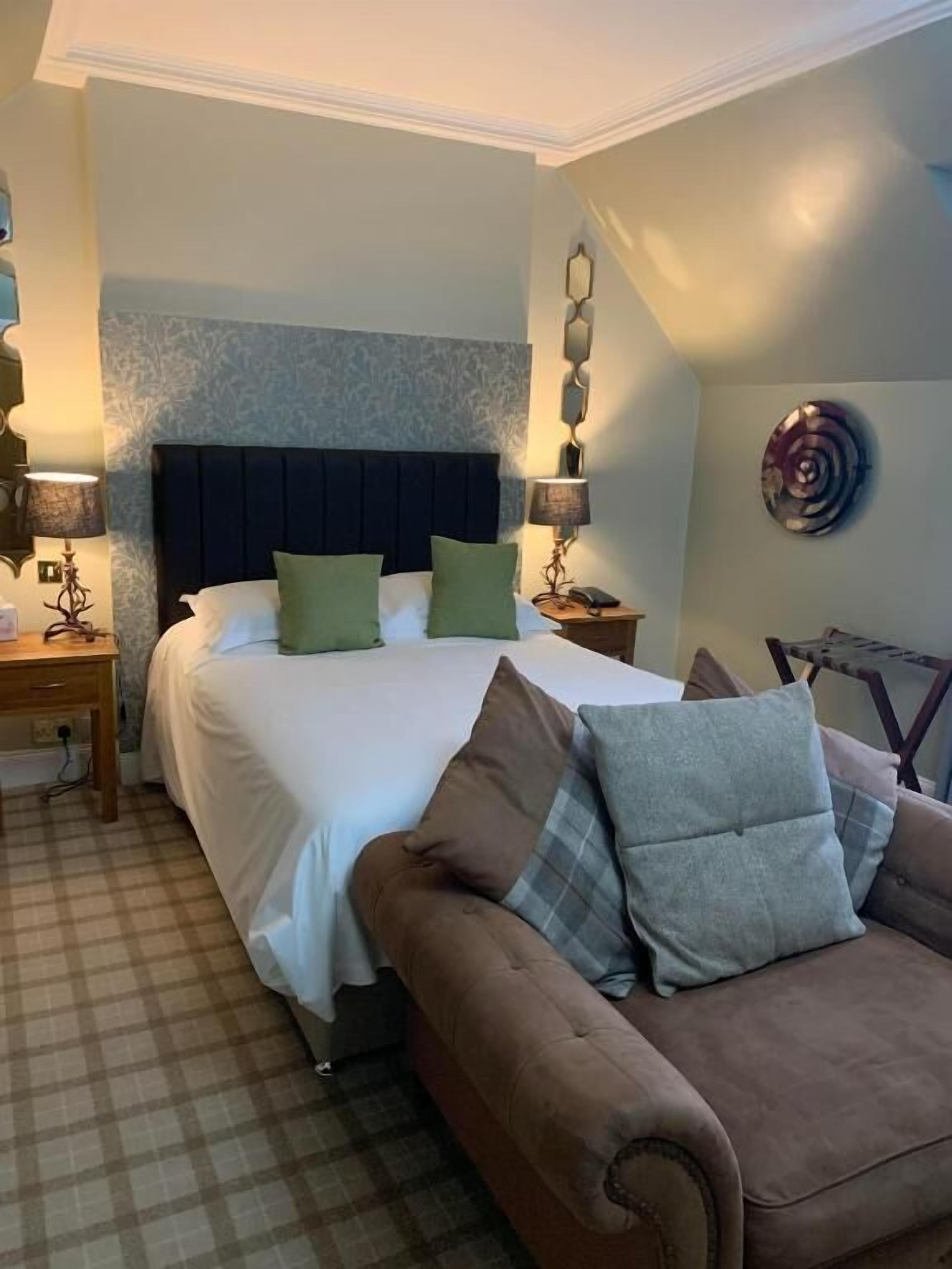 Quarto de hotel com uma cama, um sofá marrom, uma mesa de cabeceira, um abajur e um objeto decorativo na parede.