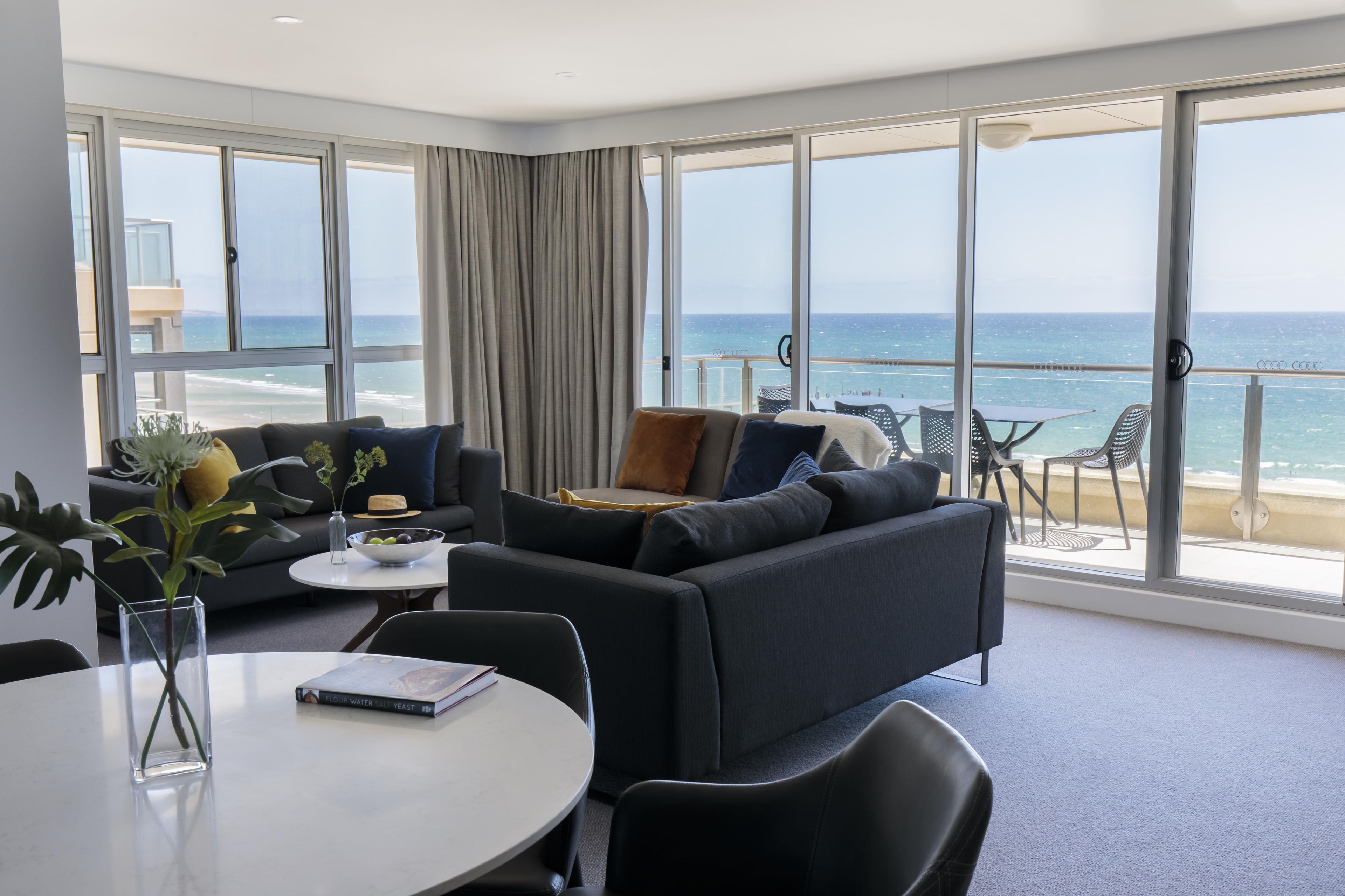 Quarto de hotel moderno com uma varanda grande, um conjunto de sofás, uma mesa de jantar e vista para o mar.