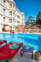 Uma área de piscina de hotel com cadeiras de sol vermelhas, uma mesa de vidro com uma garrafa e um livro, e uma piscina azul cristalina.