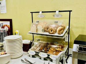 Buffet de café da manhã com seções identificadas para pães, bagels, palitos de fruta, muffins e croissants.