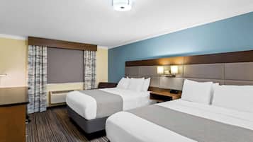 Quarto de hotel com duas camas, uma escrivaninha, uma cadeira, uma janela com cortinas, uma luminária na parede e uma luz no teto.