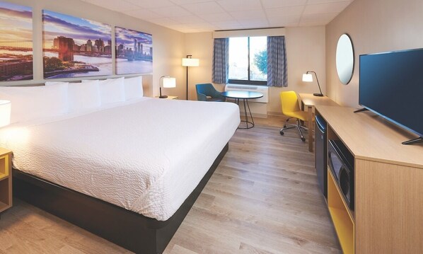 Quarto de hotel com uma cama grande, uma escrivaninha com televisão de tela plana, uma cadeira, uma mesinha e uma janela com cortinas.