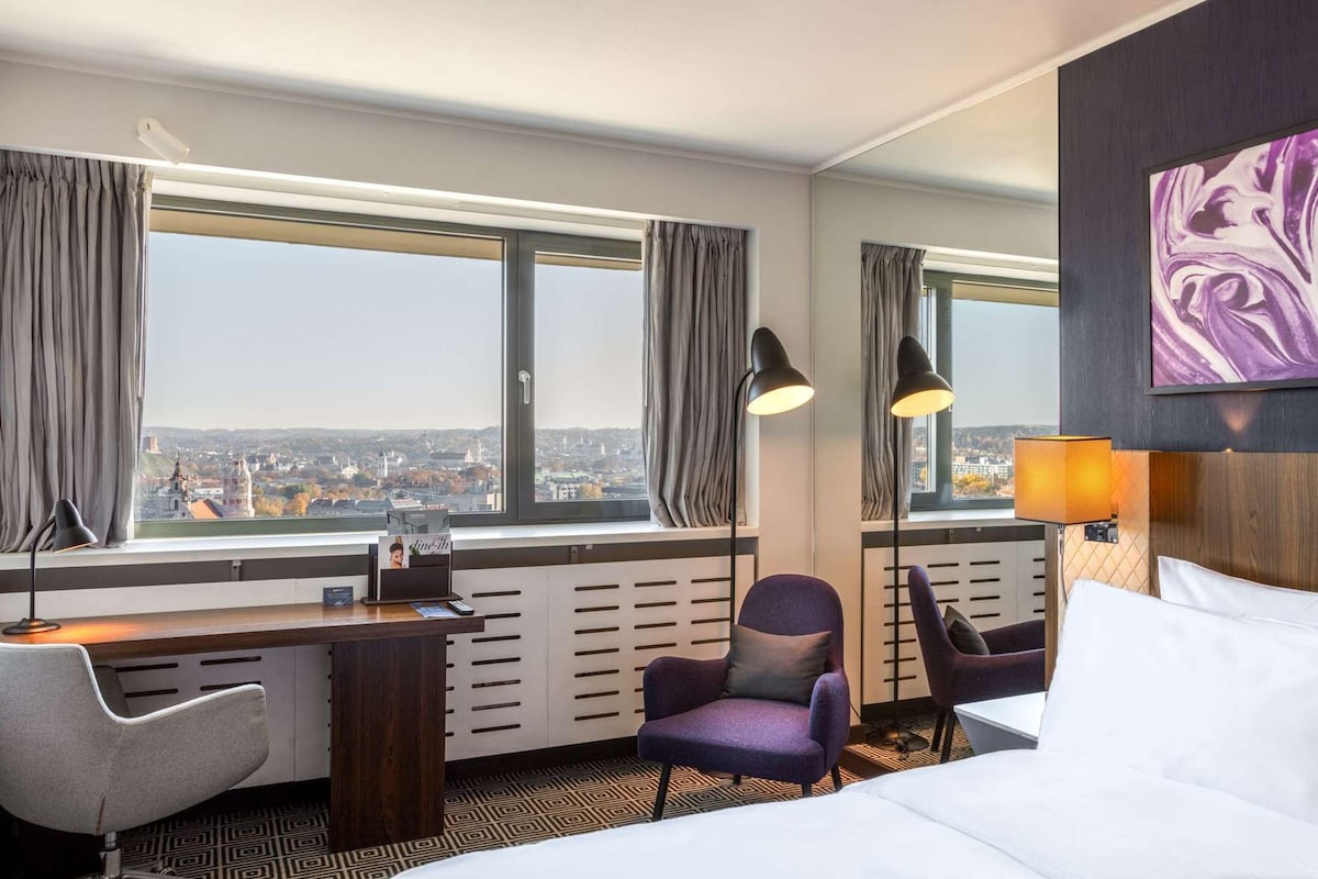 Quarto de hotel com uma escrivaninha, duas cadeiras, uma cama e uma janela grande com vista para a cidade.