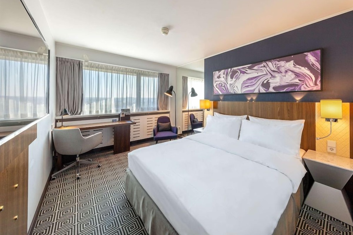 Quarto de hotel com uma cama grande, uma escrivaninha, uma cadeira e uma janela ampla com cortinas.