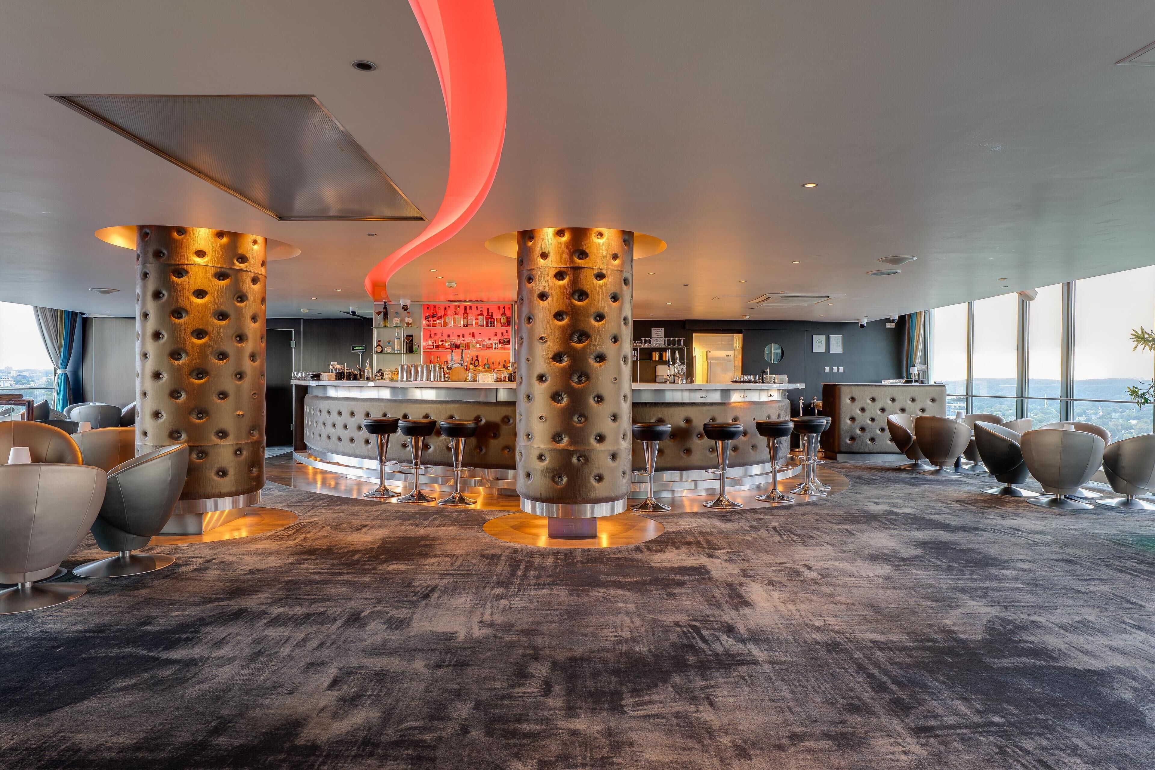 Uma área de bar moderna com uma mesa redonda central, cadeiras de encosto alto e colunas decorativas.