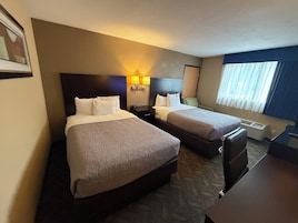 Quarto de hotel com duas camas, uma escrivaninha, uma cadeira, janela com cortinas e luminária na parede.