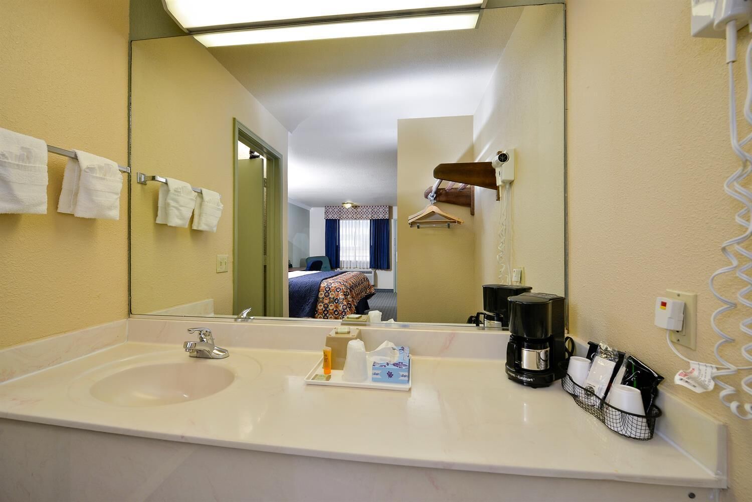 Uma casa de banho de hotel com um espelho grande, lavatório, máquina de café e cabideiro.